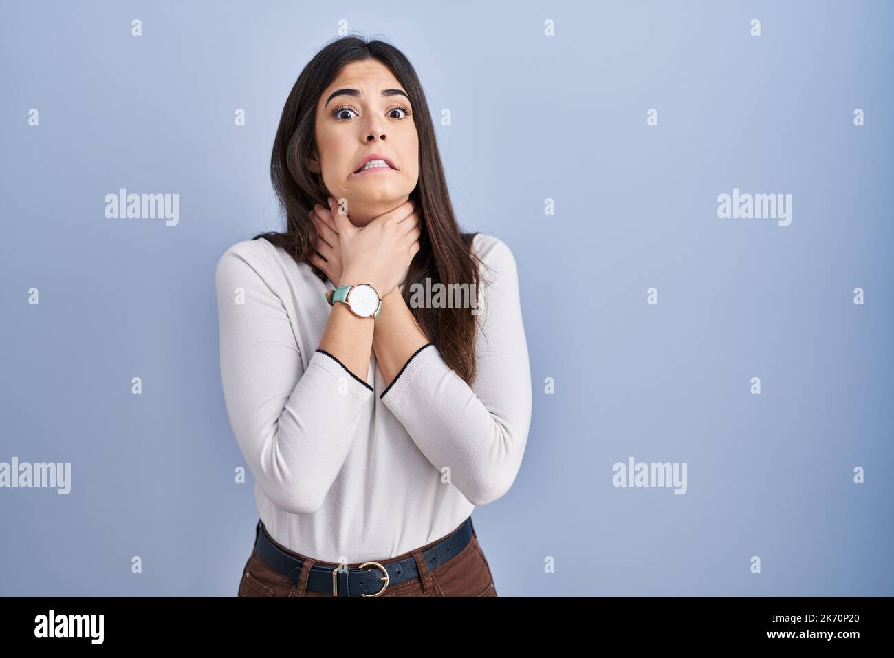 Young brunette woman standing over blue background shouting suffocate ...