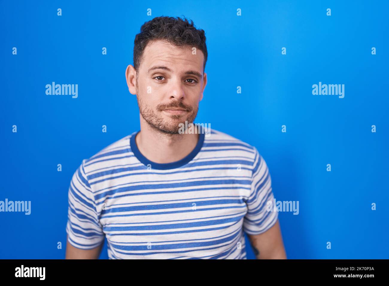 Young hispanic man standing over blue background looking sleepy and ...