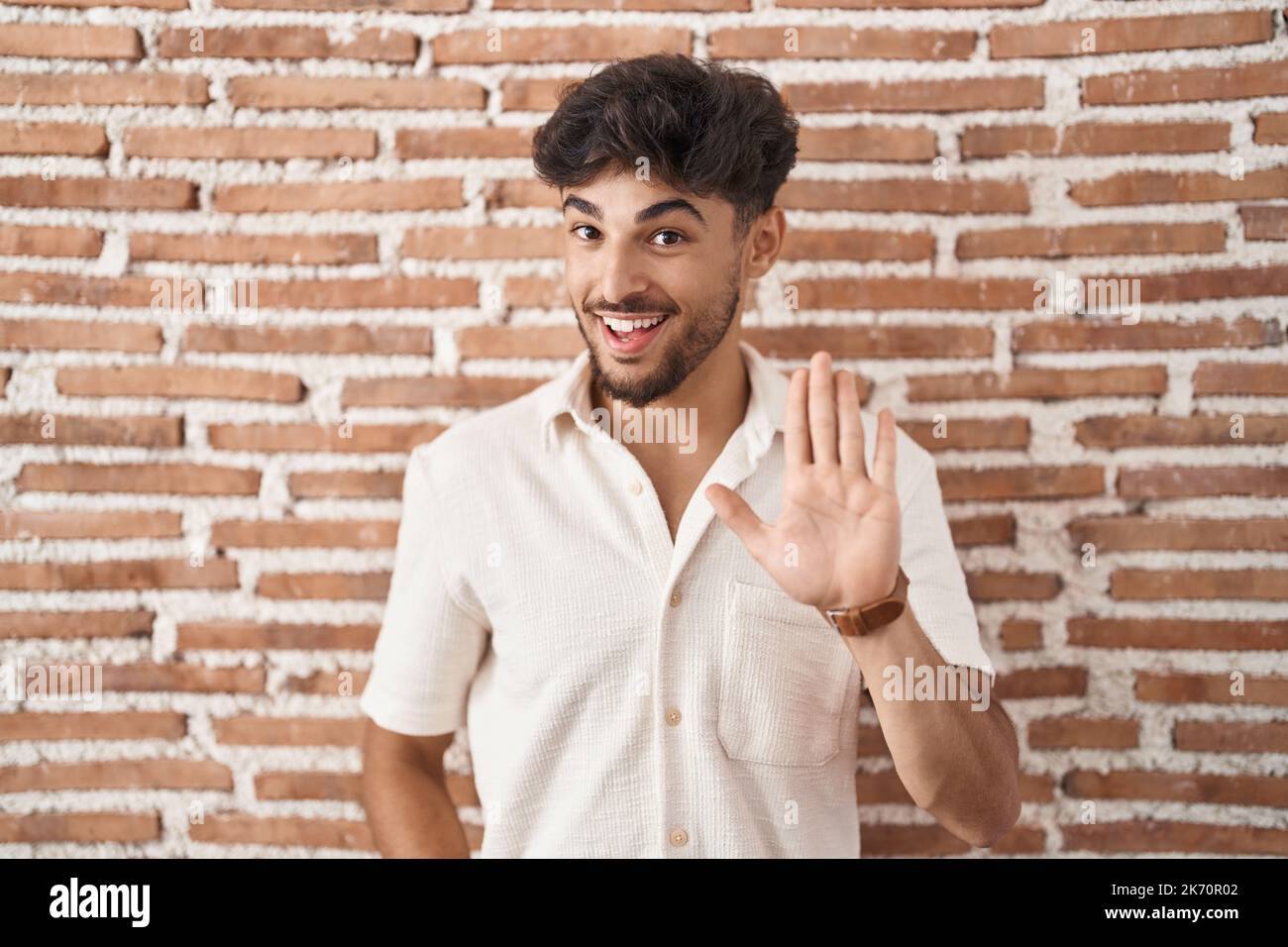 Arab man with beard standing over bricks wall background waiving saying ...