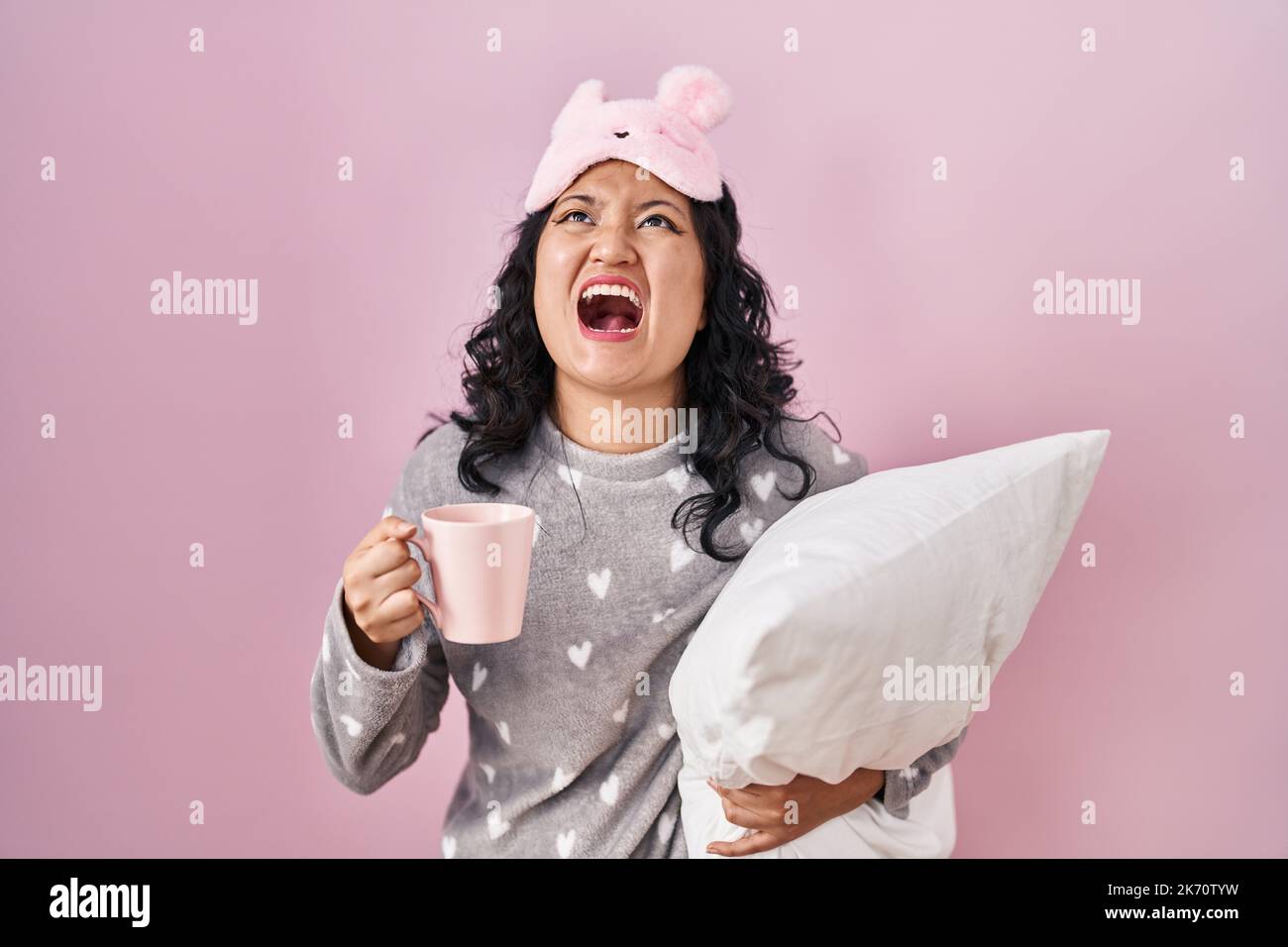 Young asian woman wearing sleep mask and pajama hugging pillow drinking ...