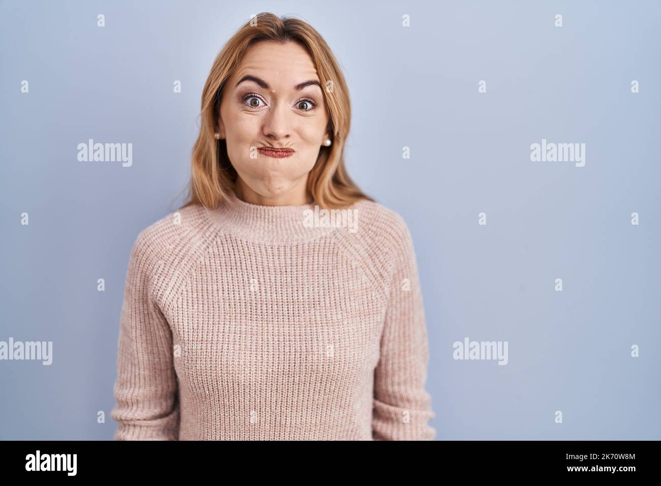 Hispanic woman standing over blue background puffing cheeks with funny ...