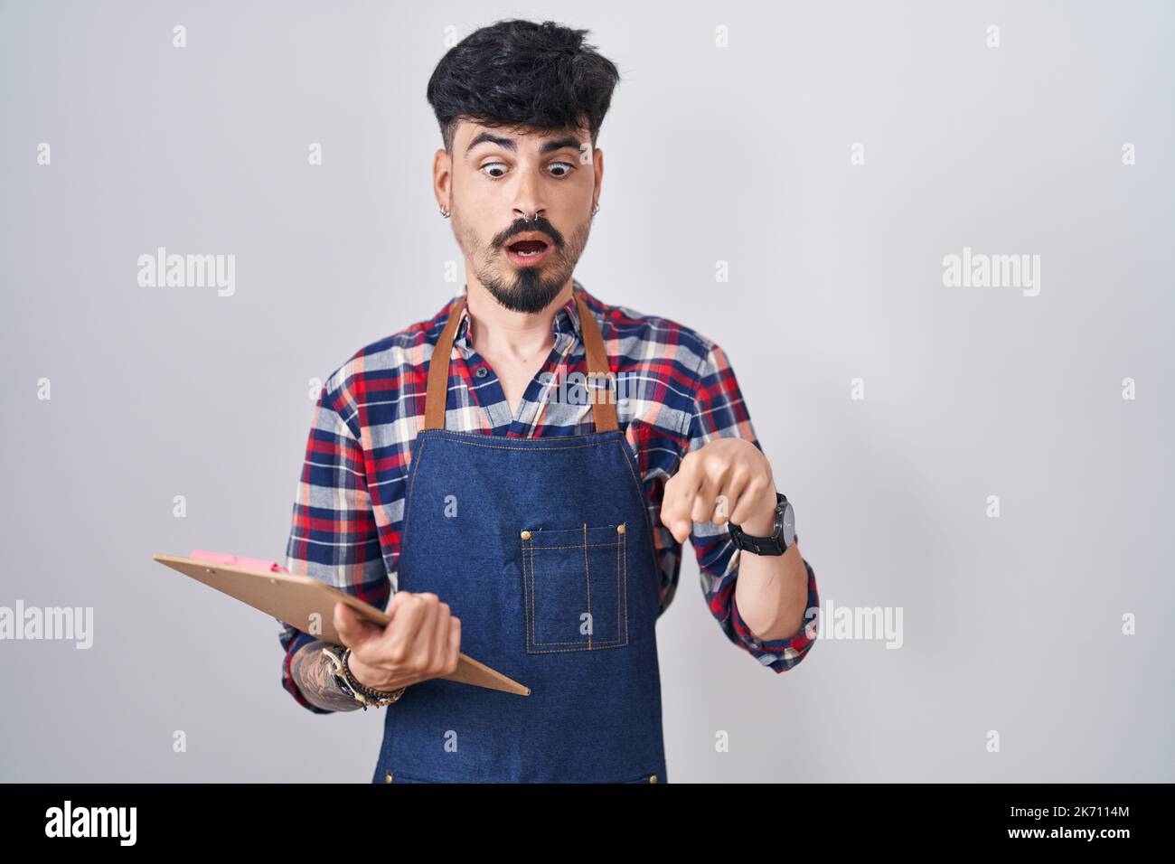 Young hispanic man with beard wearing waiter apron holding clipboard ...
