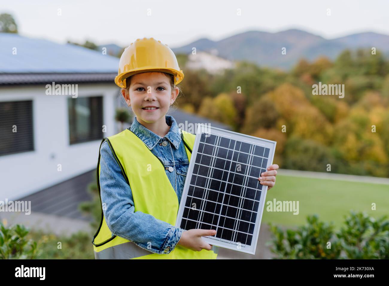 Little girl with protective helmet and reflective vest holding photovoltaics solar panel ...