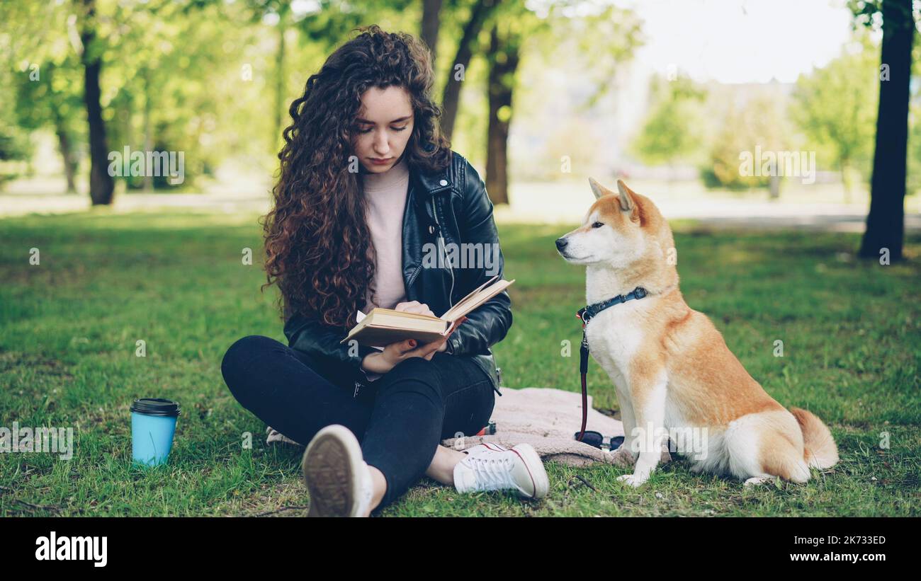 Pretty young lady is reading book in the park and patting cute dog ...