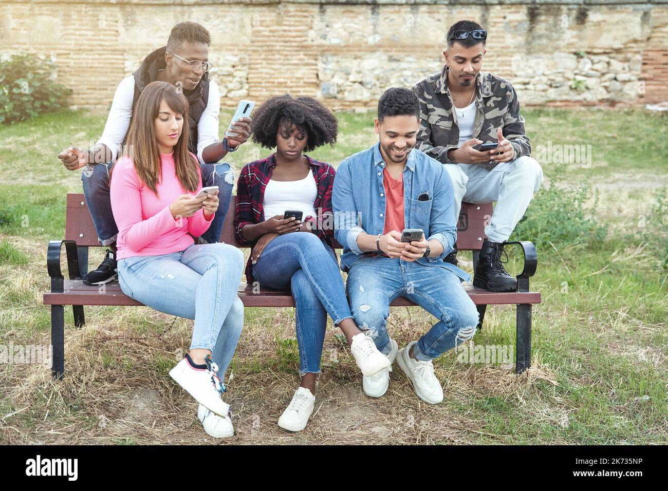 Group of multi-ethnic friends using their mobile phones while sitting on a bench together ...