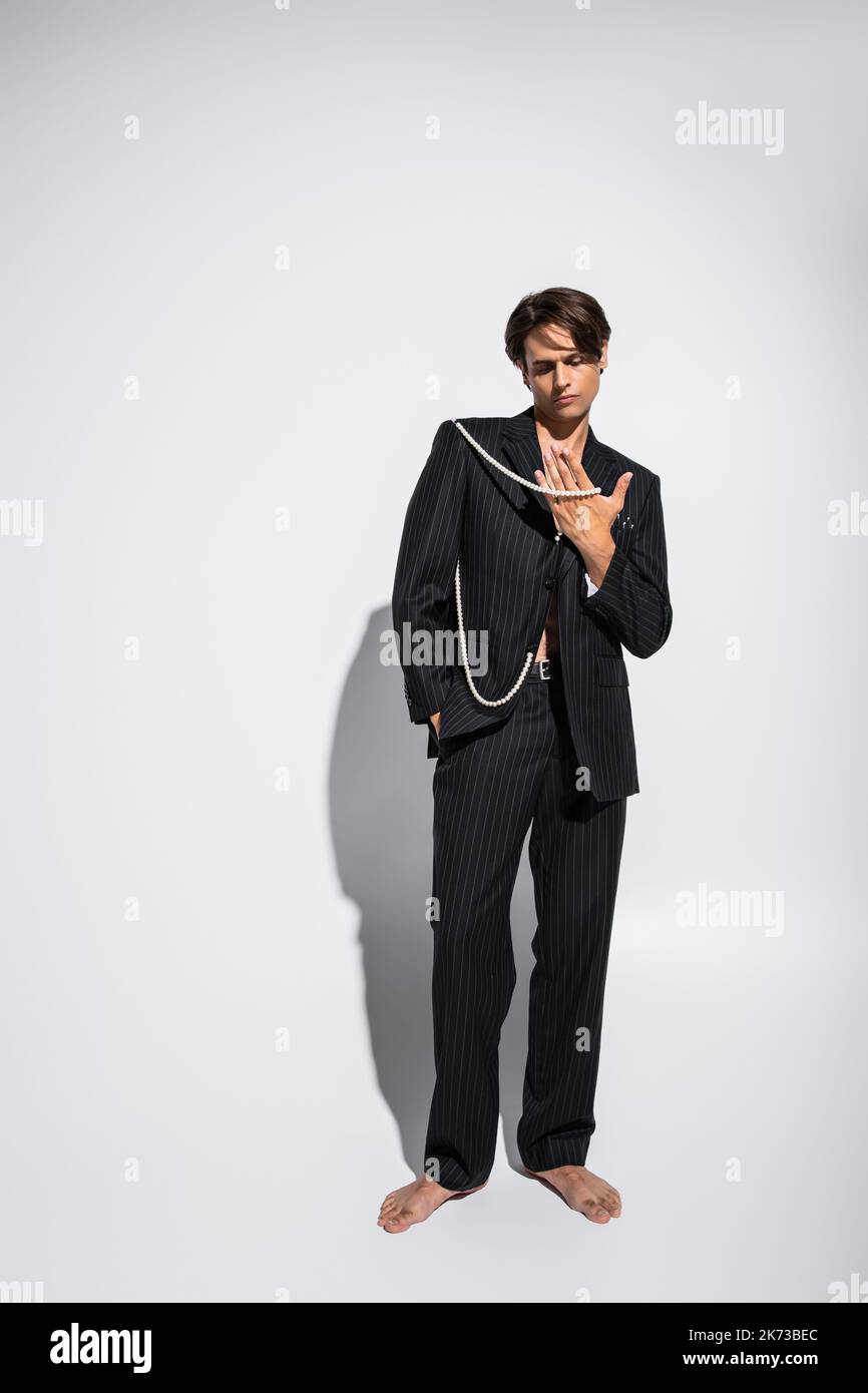 full length of barefoot and brunette man in trendy suit holding pearls ...