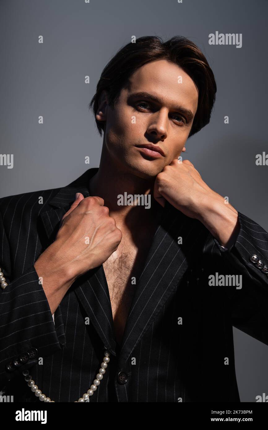 brunette man looking at camera while adjusting trendy suit on dark grey ...