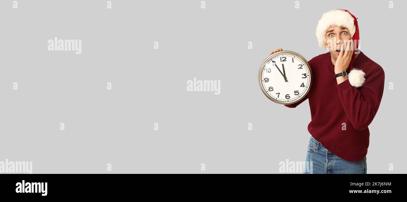 Surprised young man in Santa hat holding clock on light background with ...