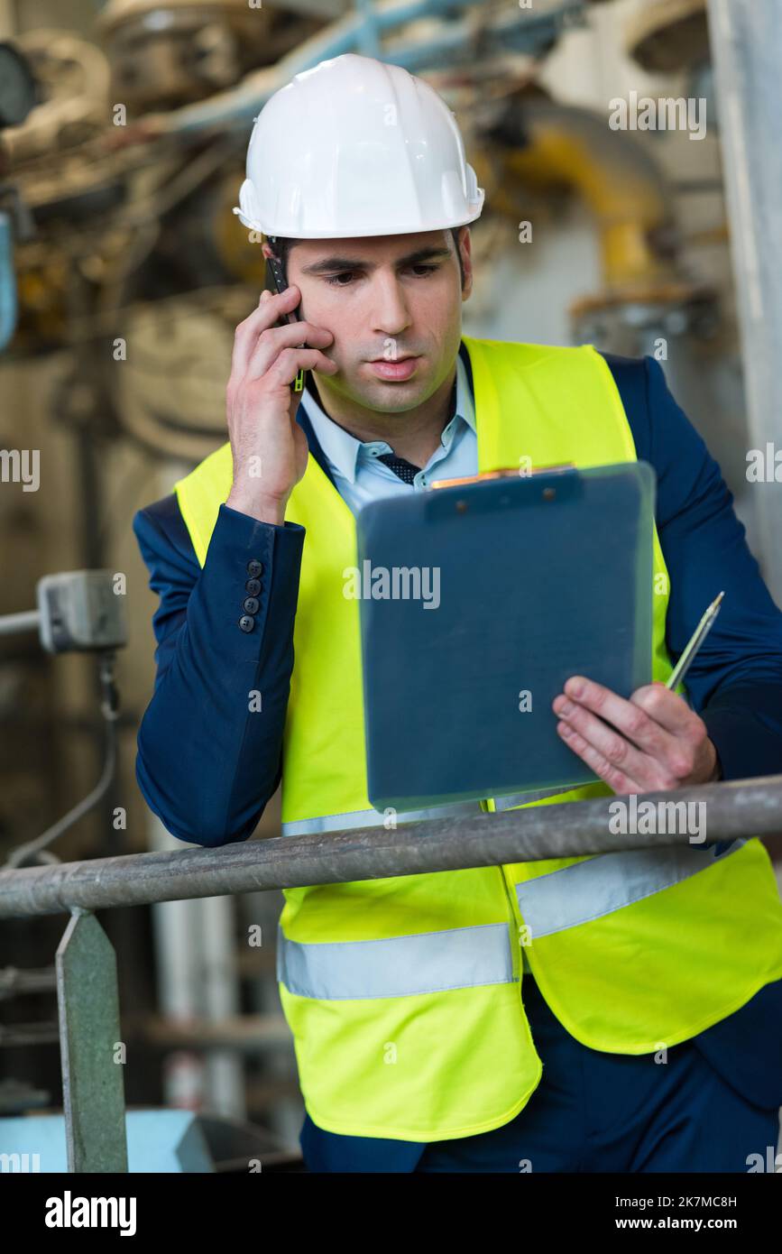 male engineer in factory holding clipboard and talking on telephone ...