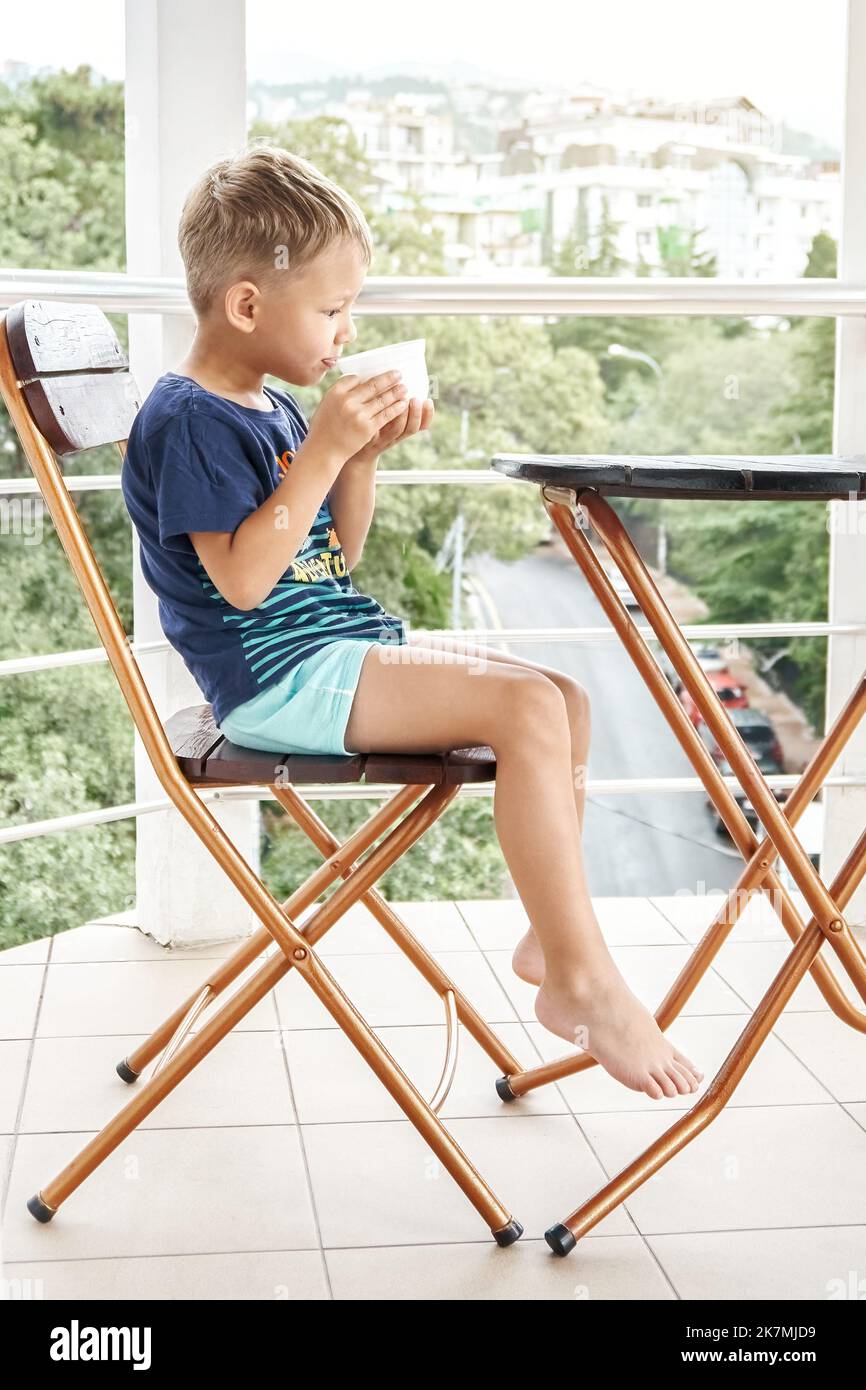 Preschooler boy drinks warm milk from mug sitting on chair in early ...