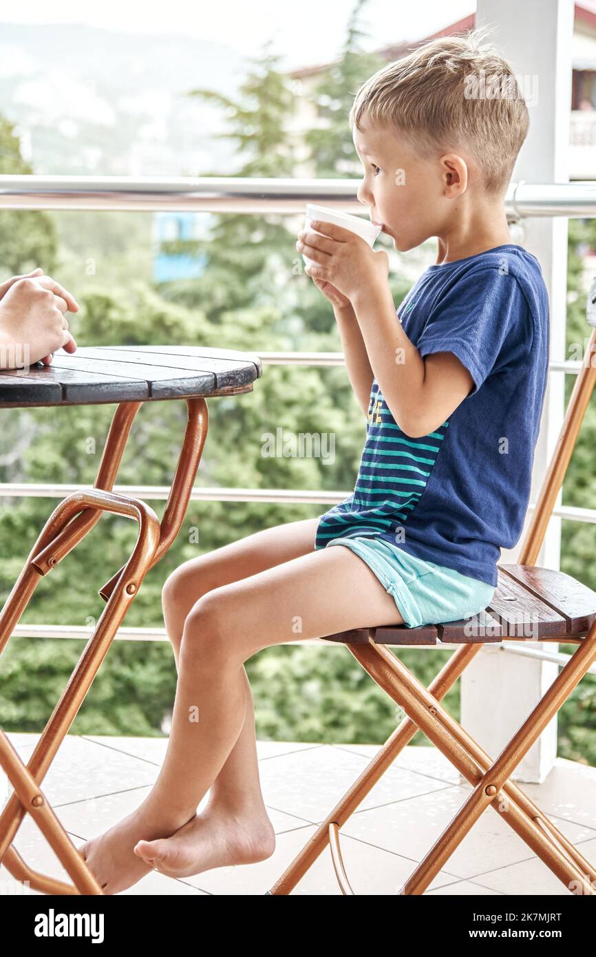 Preschooler boy drinks warm milk from mug sitting on chair in early ...