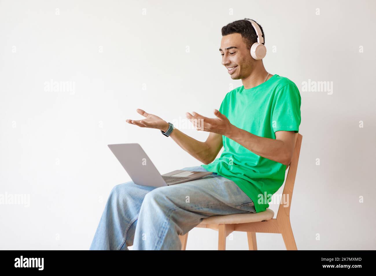 Positive arab guy sitting on chair over light wall, using laptop and ...