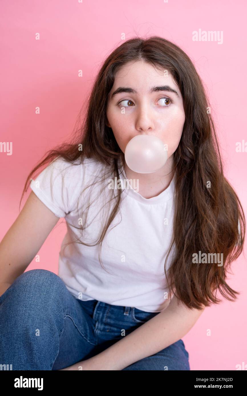 Portrait Of A Young Woman Blowing Bubble Gum Bubbles Against A Pink