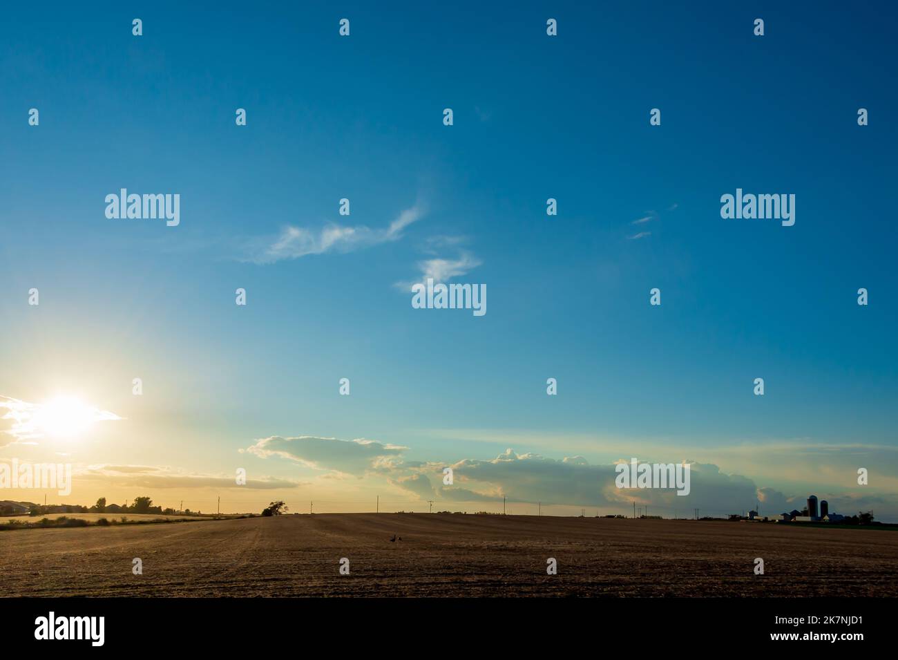 A setting sun and clouds highlight a harvested autumn farm field and ...