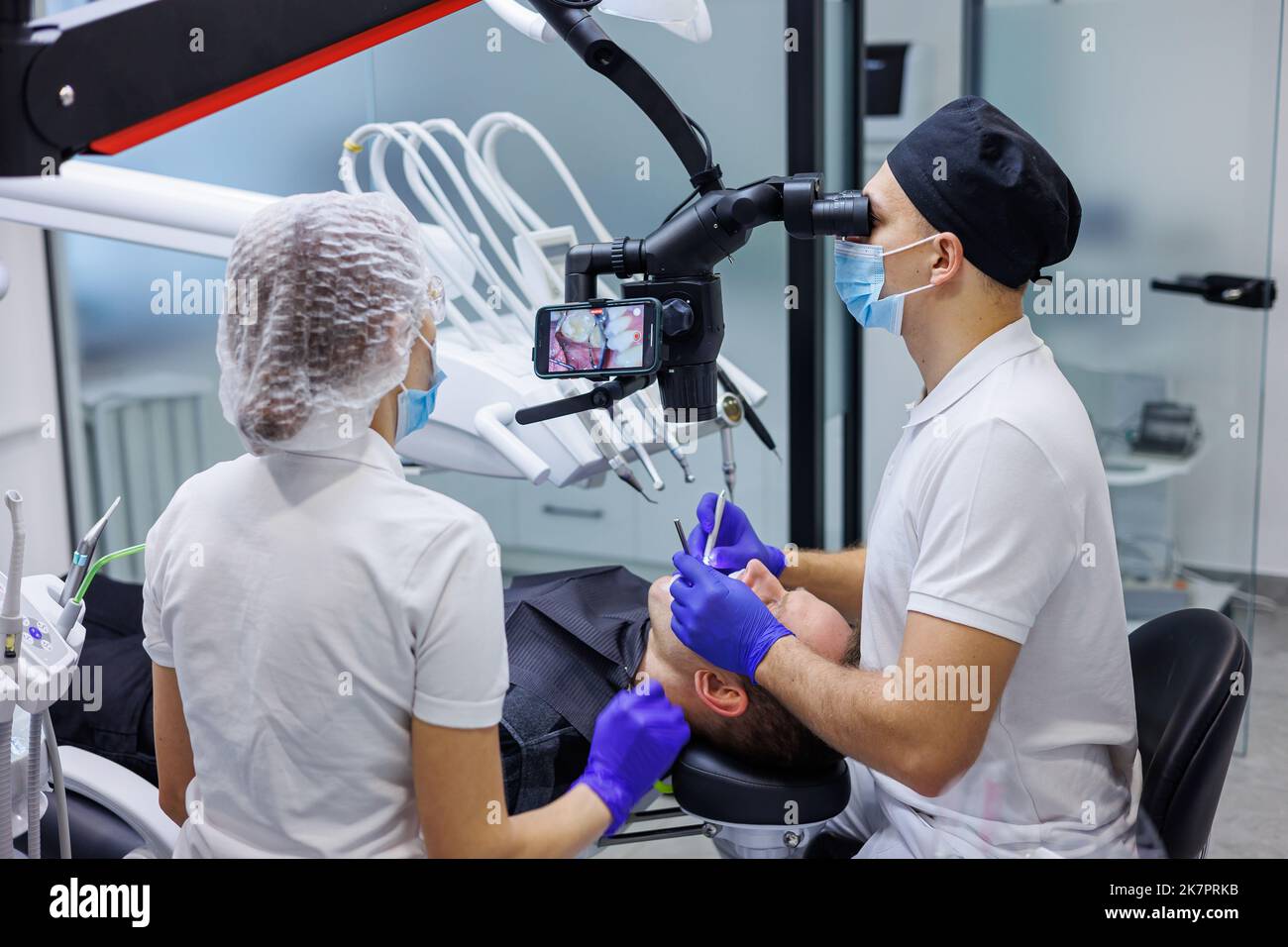 A dentist and an assistant treat a patient's teeth using a microscope ...