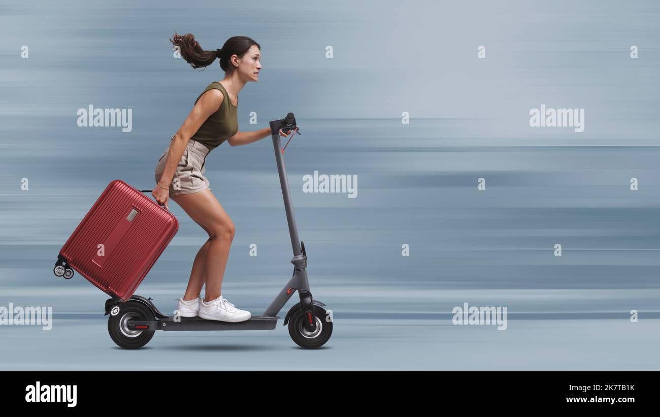 Tourist woman riding a fast electric scooter and carrying a trolley bag ...