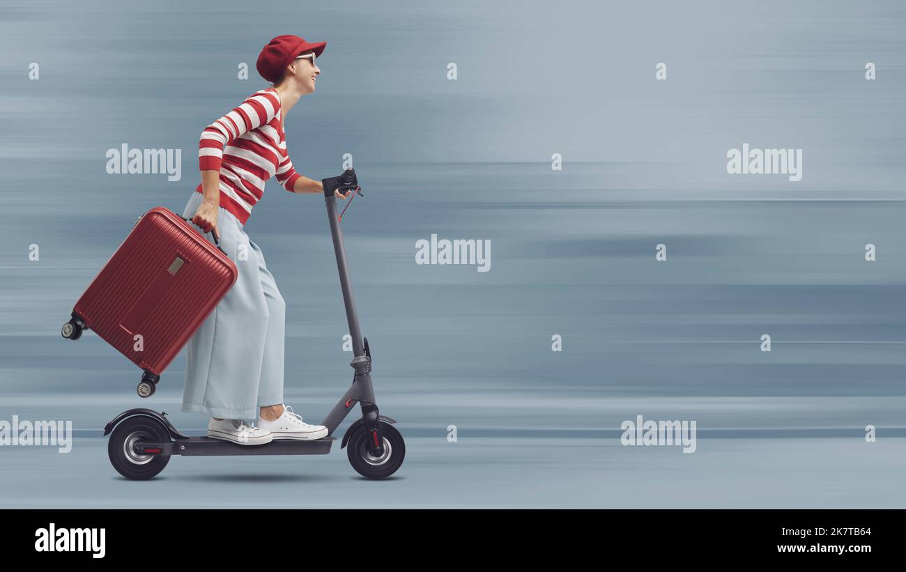 Tourist woman riding a fast electric scooter and carrying a trolley bag ...