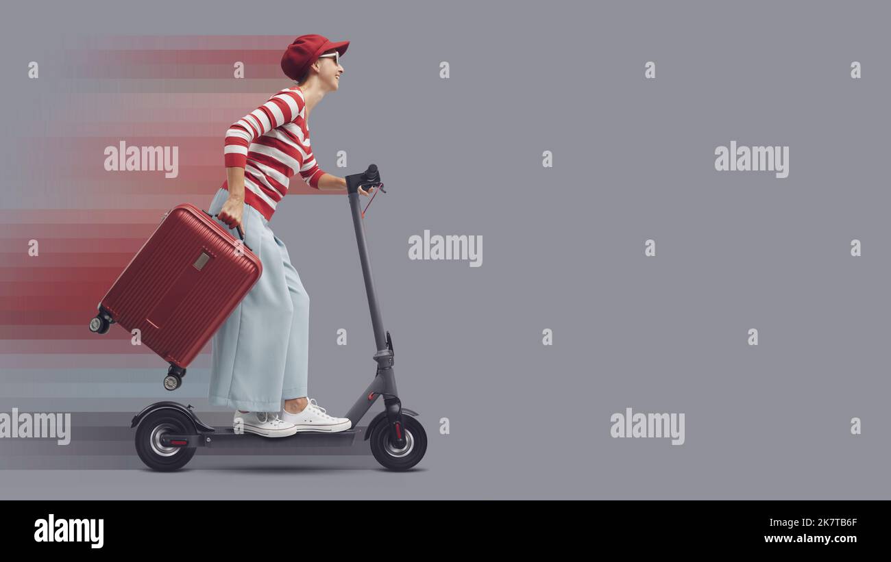 Tourist woman riding a fast electric scooter and carrying a trolley bag ...