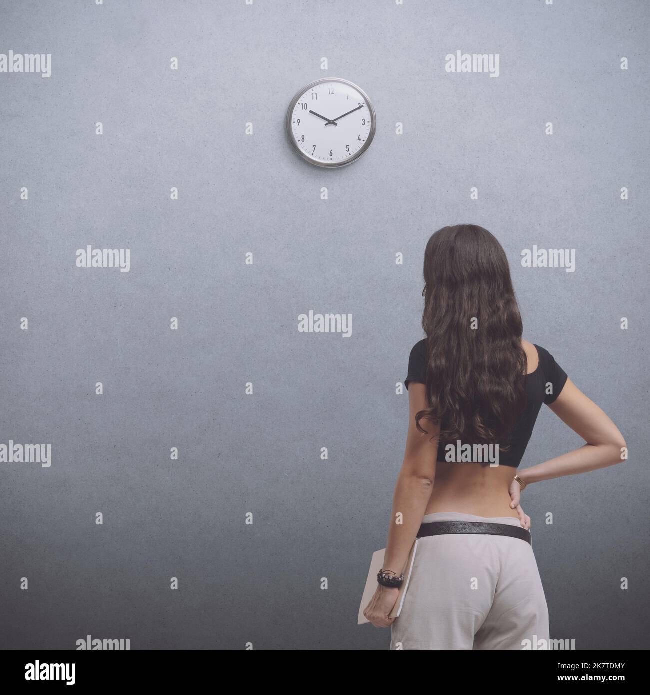 Young woman looking at the wall clock and checking the time, back view ...