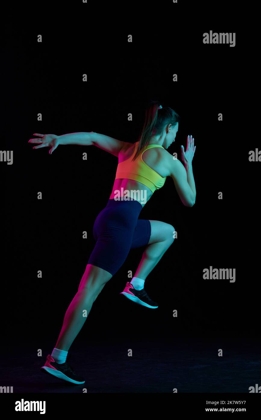 Professional female athlete running isolated on dark background in neon ...