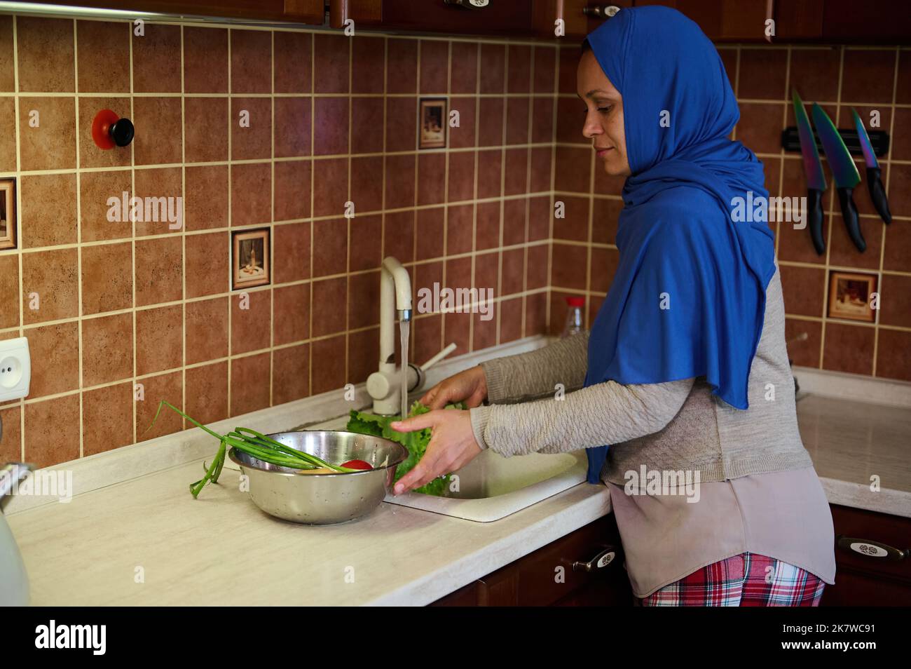 Arab Muslim woman in hijab stands by a sink in the kitchen and washes ...