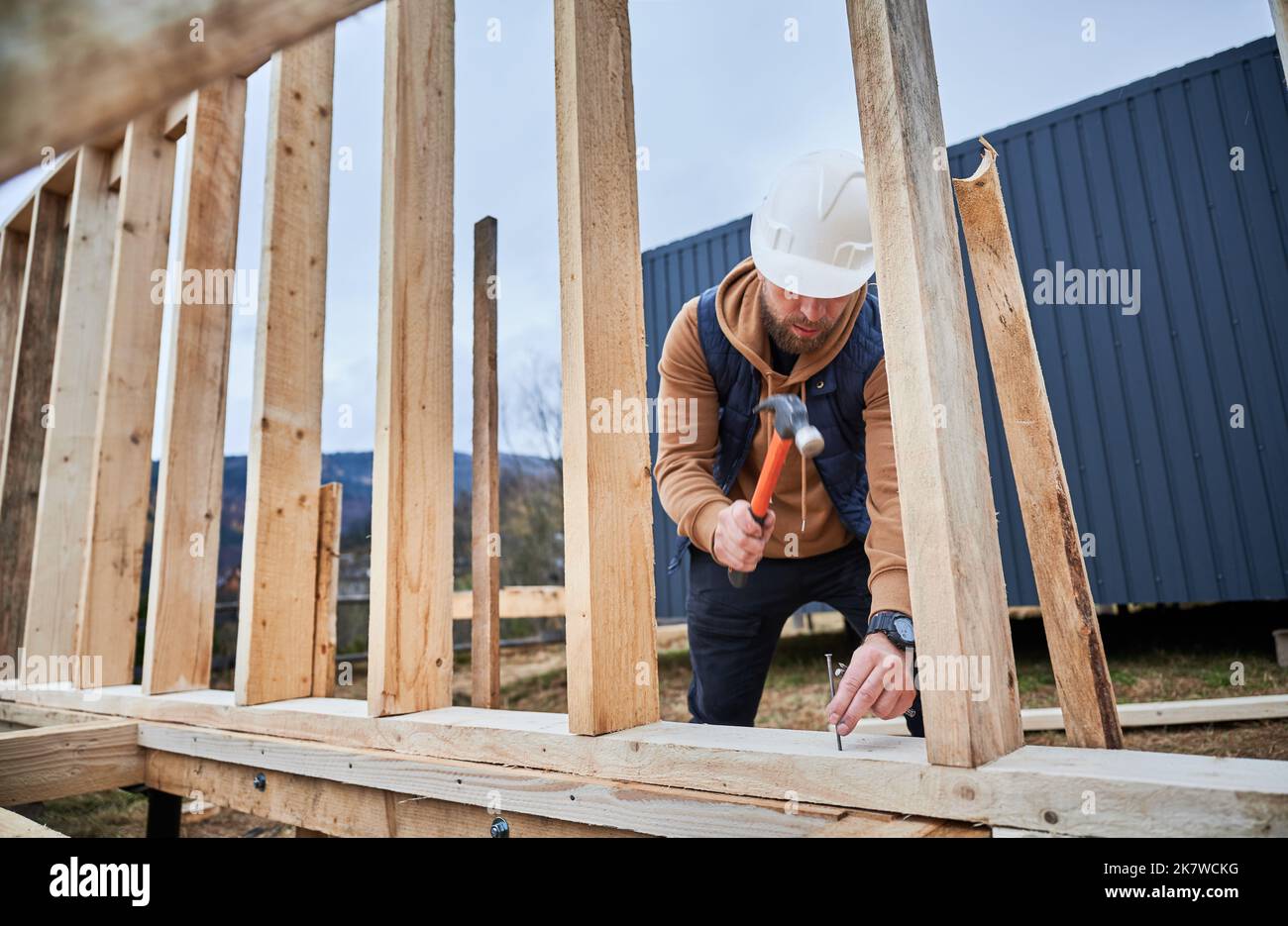 Man worker building wooden frame house on pile foundation. Carpenter hammering nail into wooden ...
