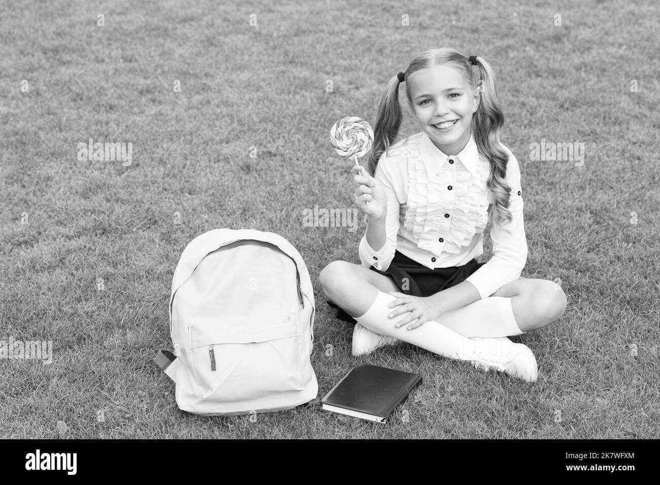 Happy girl child in school uniform hold sweet lollipop sitting on green ...