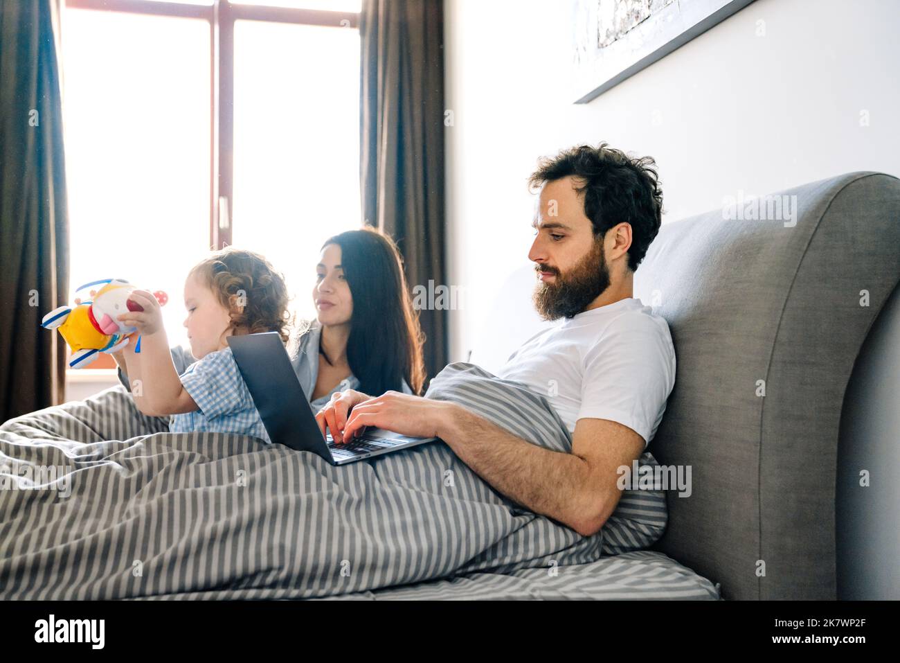 Happy family relaxing together in bed in the morning father working on ...