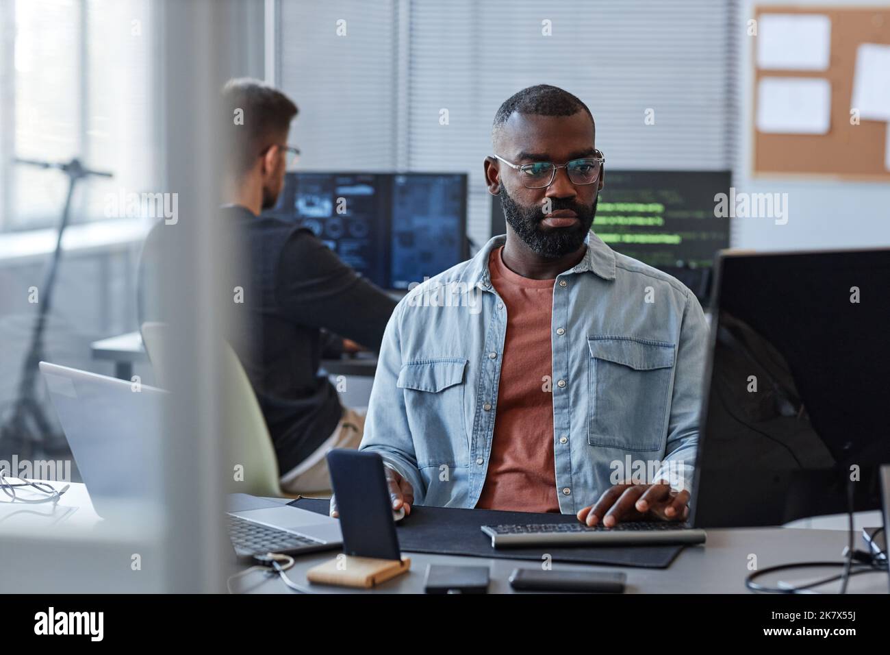 Portrait of black man using computer while programming mobile software in office behind glass ...
