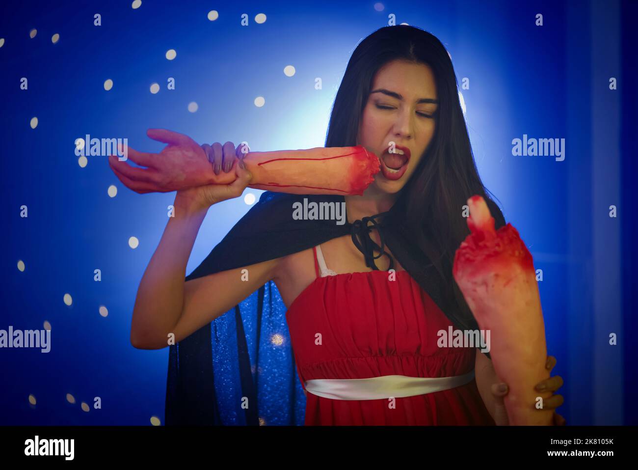 Young woman in red dress and black coat biting fake bloody arms ...