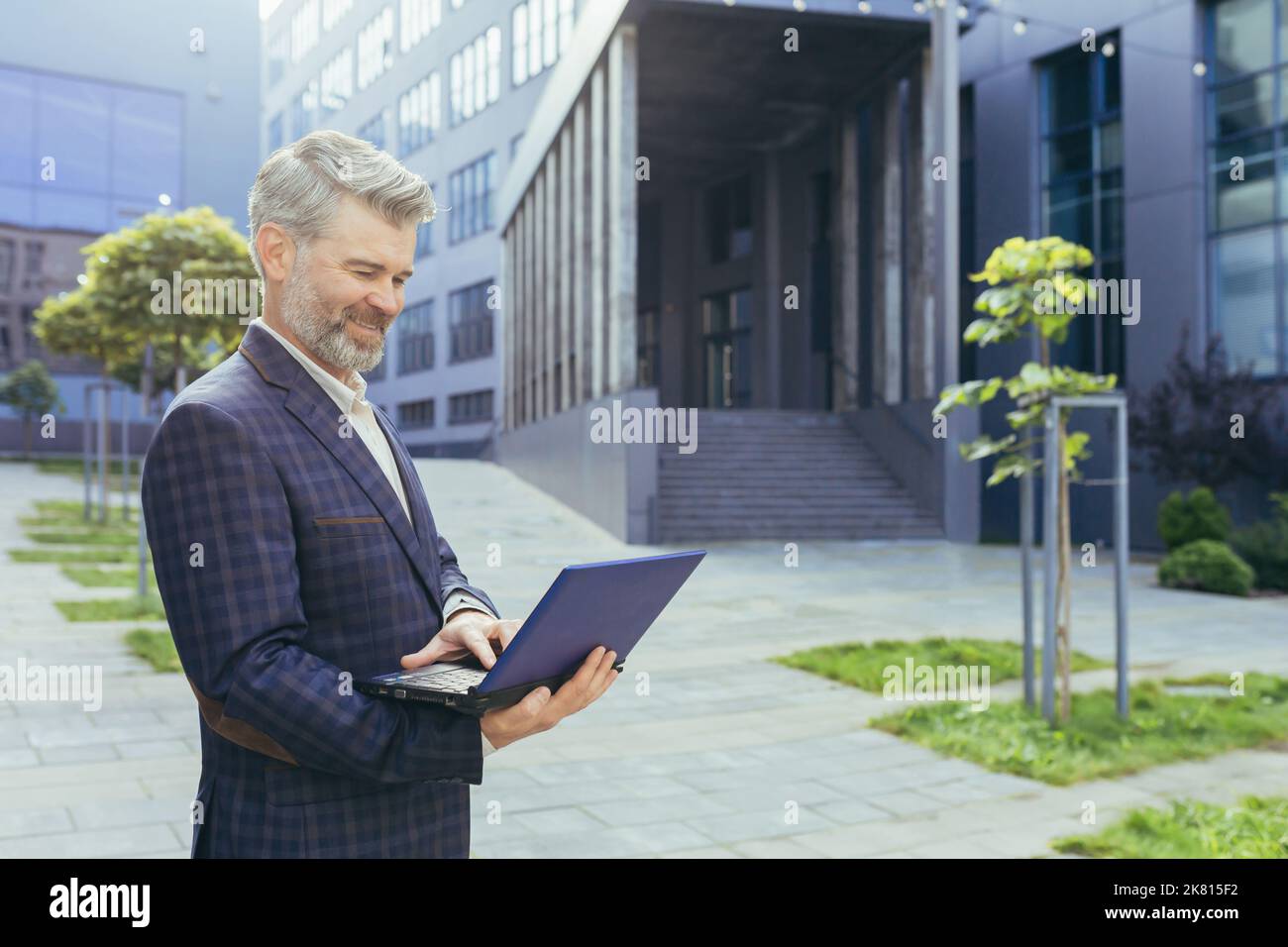 Happy and smiling boss using laptop while standing outside modern ...