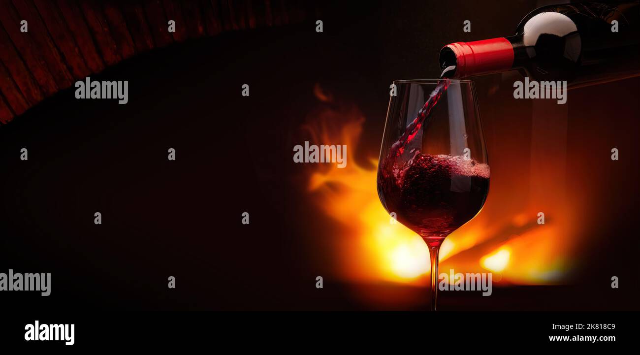 Pouring red wine into a glass at night near fireplace flames. Cozy ...