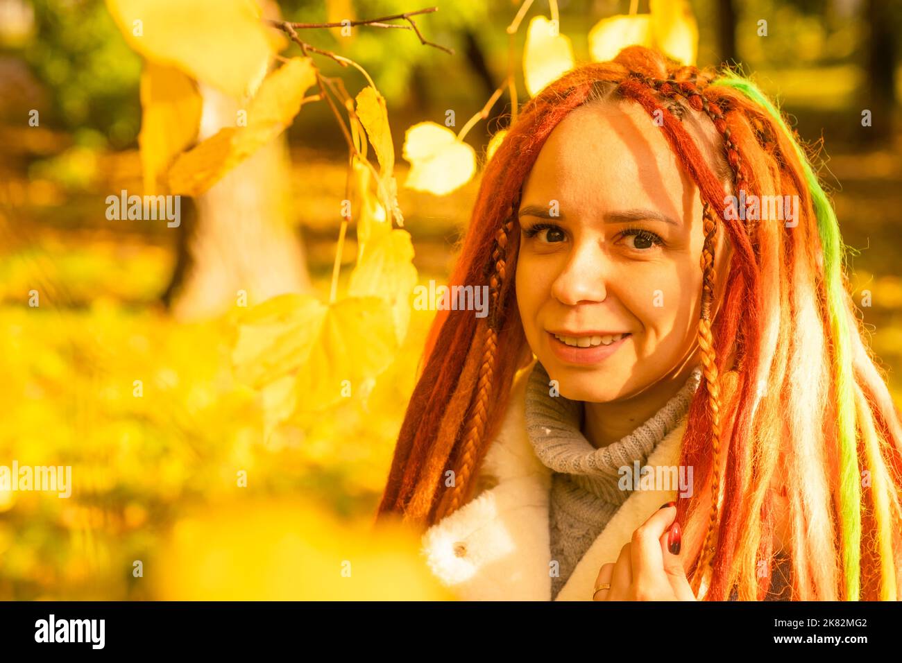 Portrait of young positive woman with ginger dreadlocks standing among ...