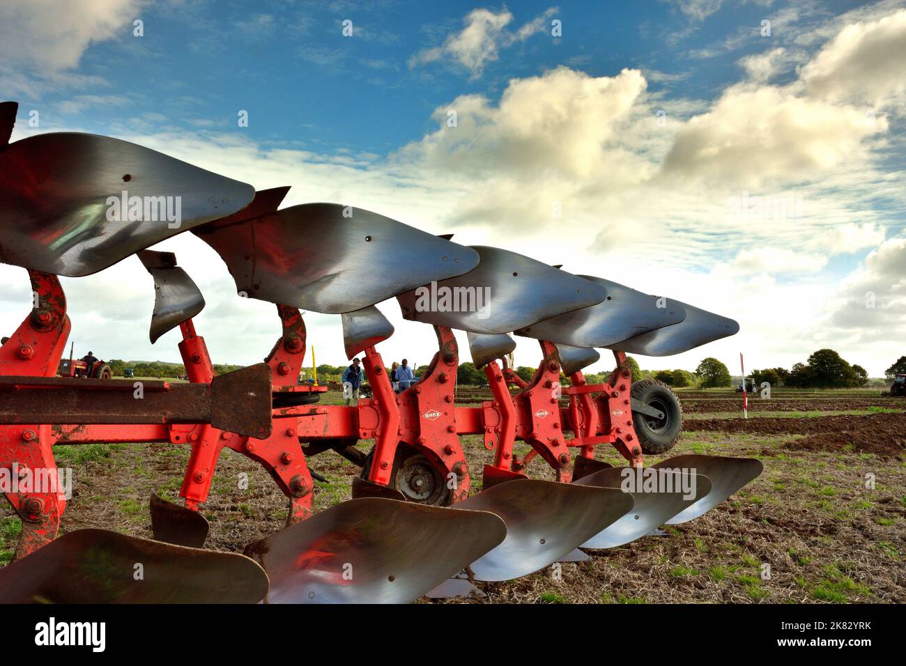 Red and silver plough in a large plowed field with blue sky and clouds ...