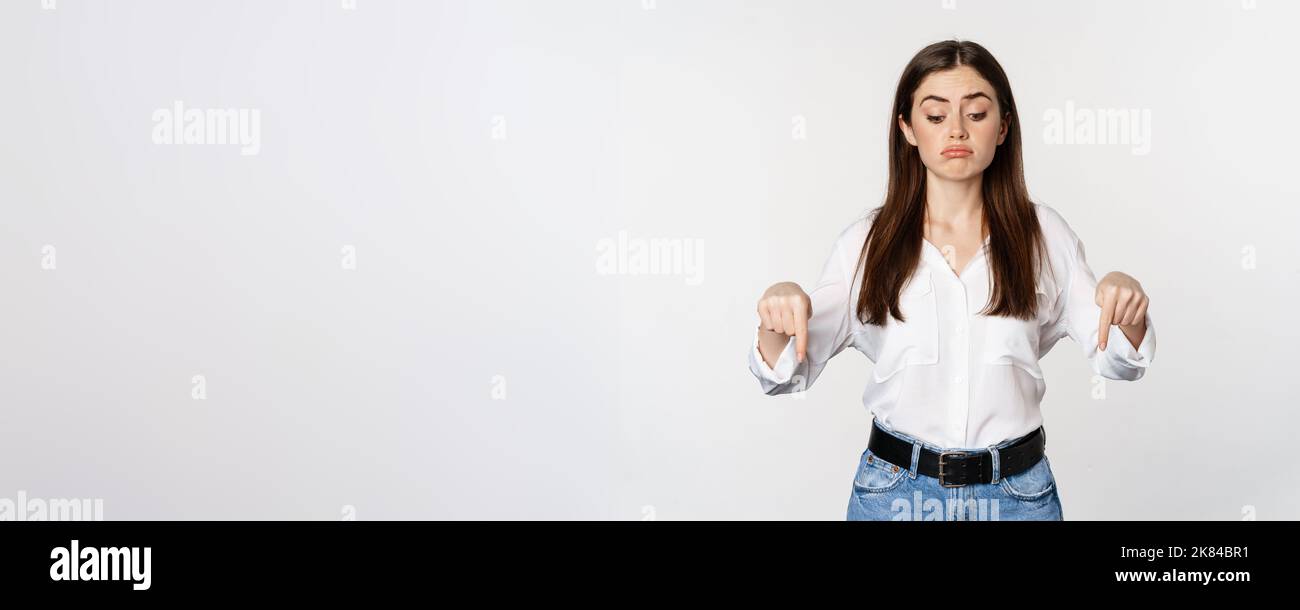 Disappointed young woman pointing fingers down, looking below with ...