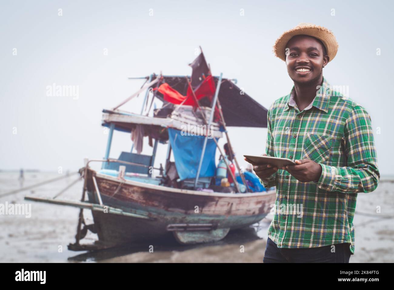 African trawler captain stands in front of the ship using a tablet to ...