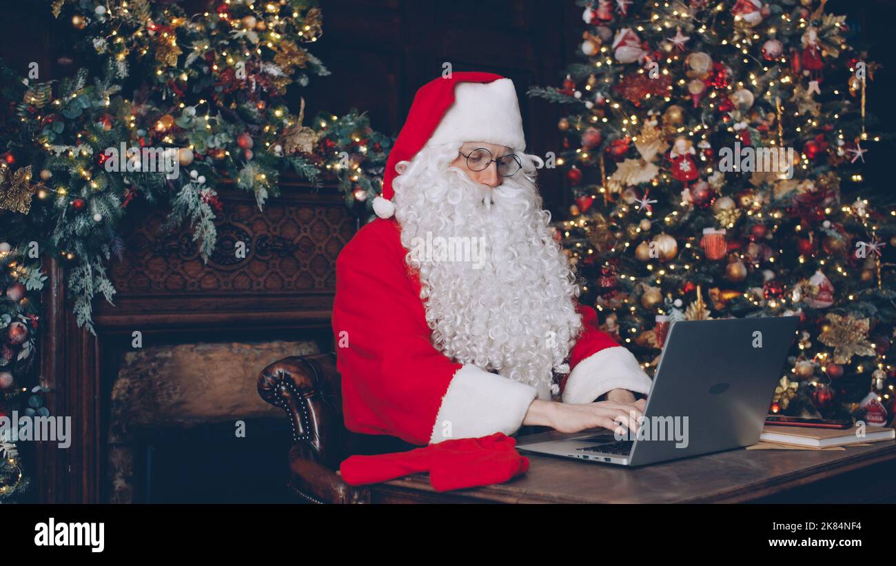 Santa in costume and glasses is typing letter with laptop computer in ...