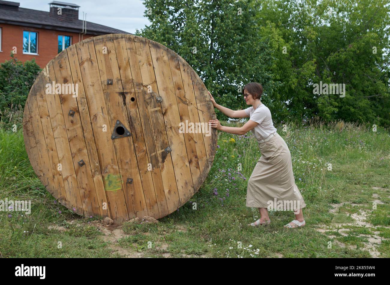 A woman in casual clothes rolls a huge wooden spool of wire Stock Photo ...