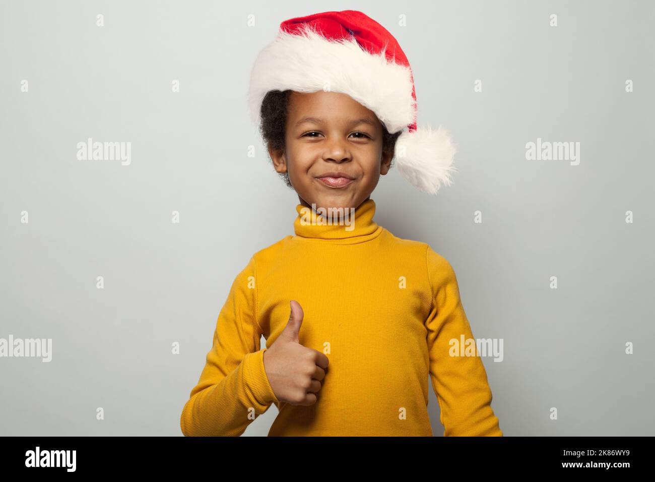 Small optimistic black child boy in Santa hat smiling and showing thumb ...