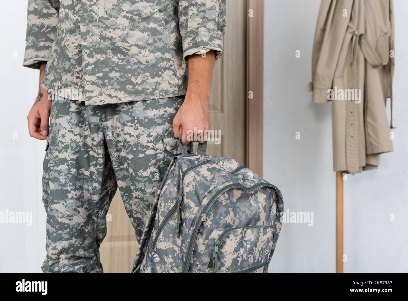 cropped view of soldier in camouflage standing with backpack at home ...