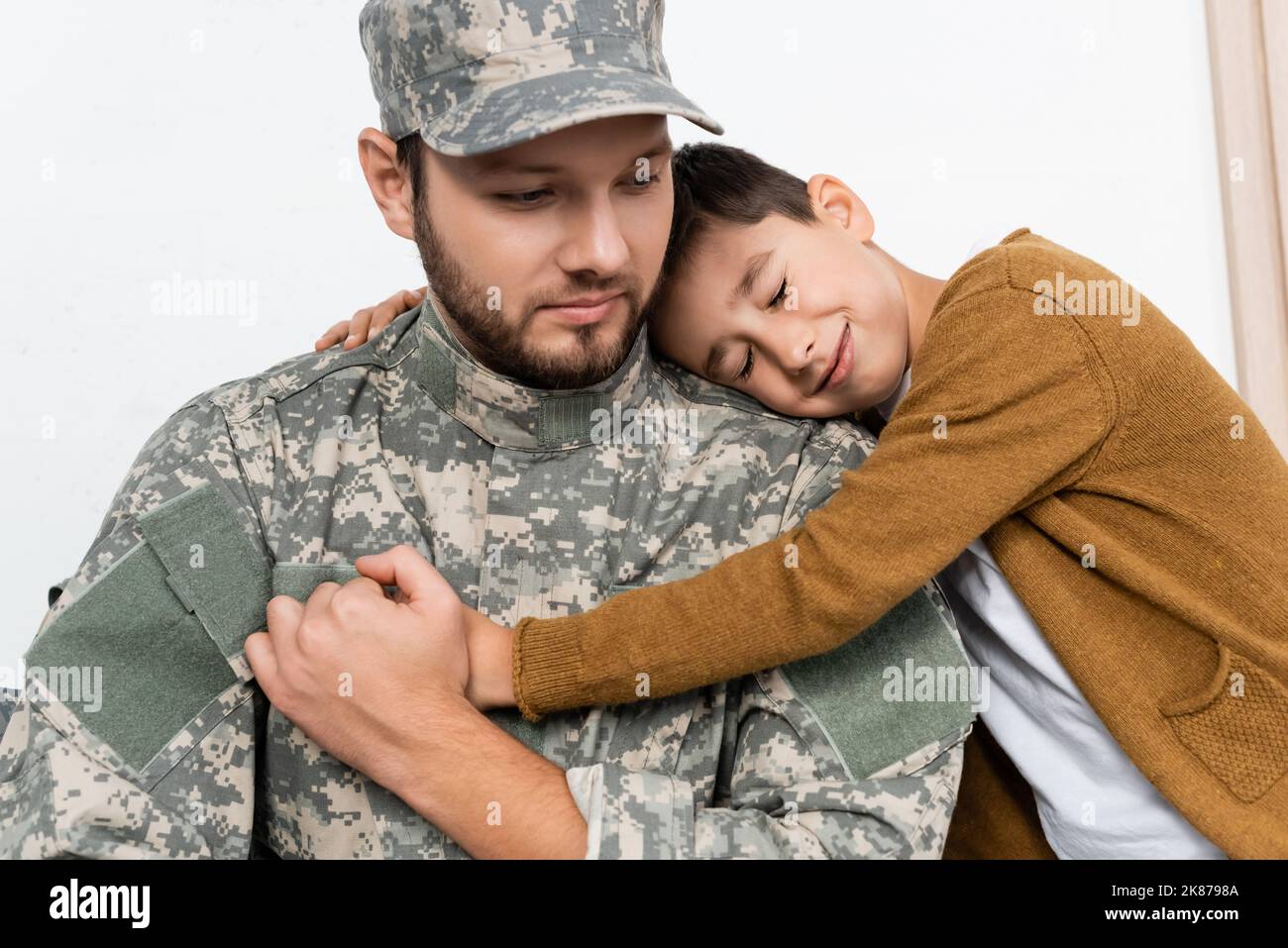 smiling kid with closed eyes hugging dad in military uniform while meeting him at home Stock ...