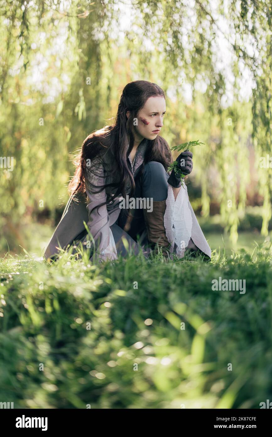 Young sad woman in image of ancient warrior sits on grass under tree in ...