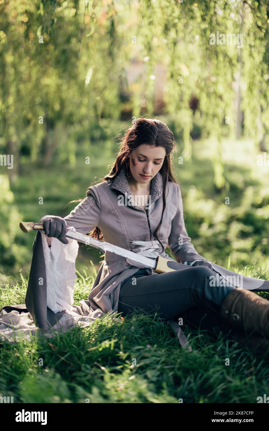 Young woman in image of ancient warrior sits and rests on grass under ...