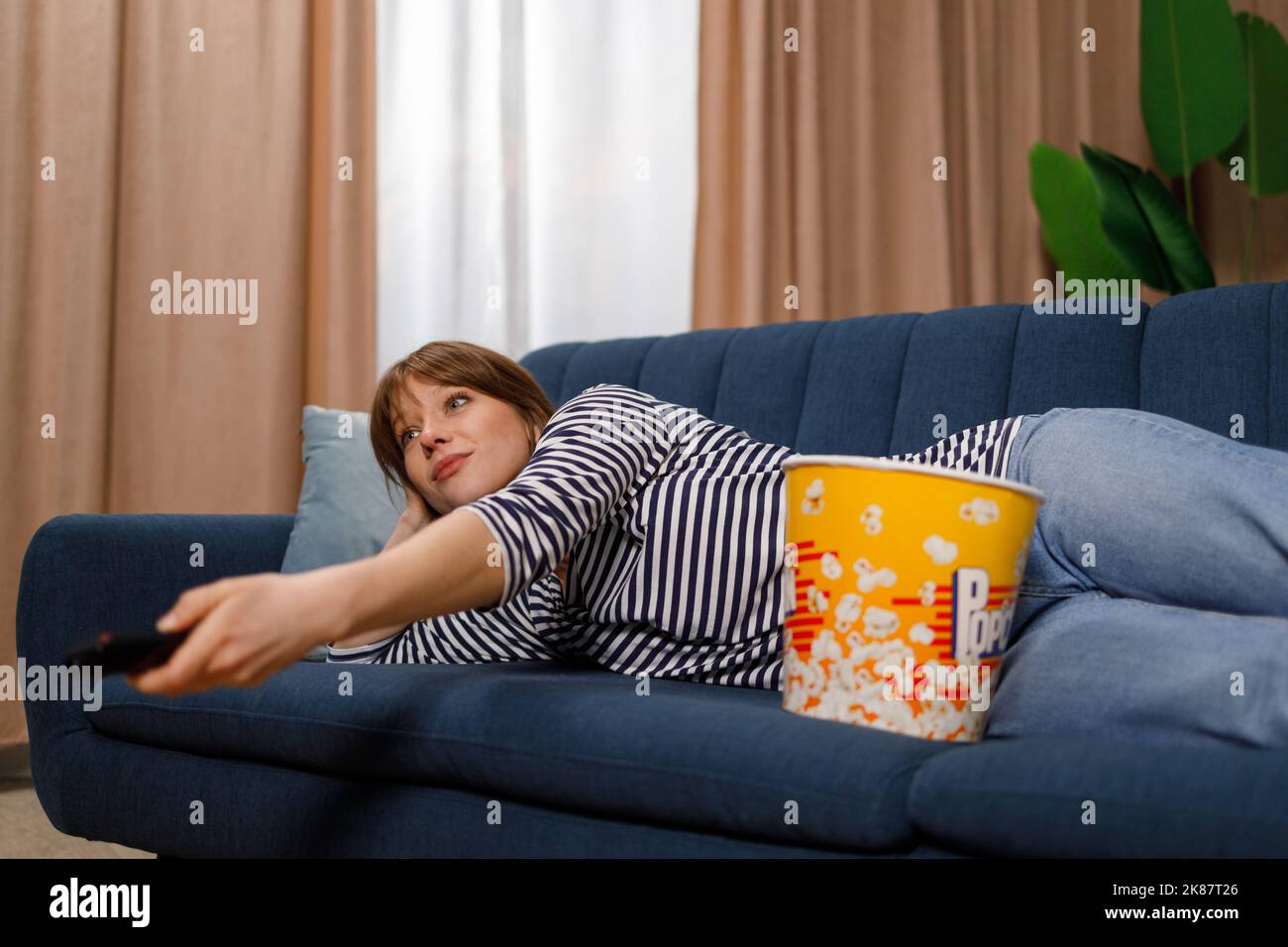 Cheerful young woman with remote control relaxing on the sofa at home ...