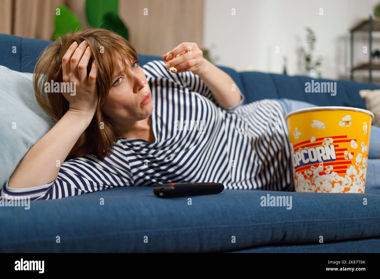 Depressed young woman lying on sofa eating popcorn and watching TV ...