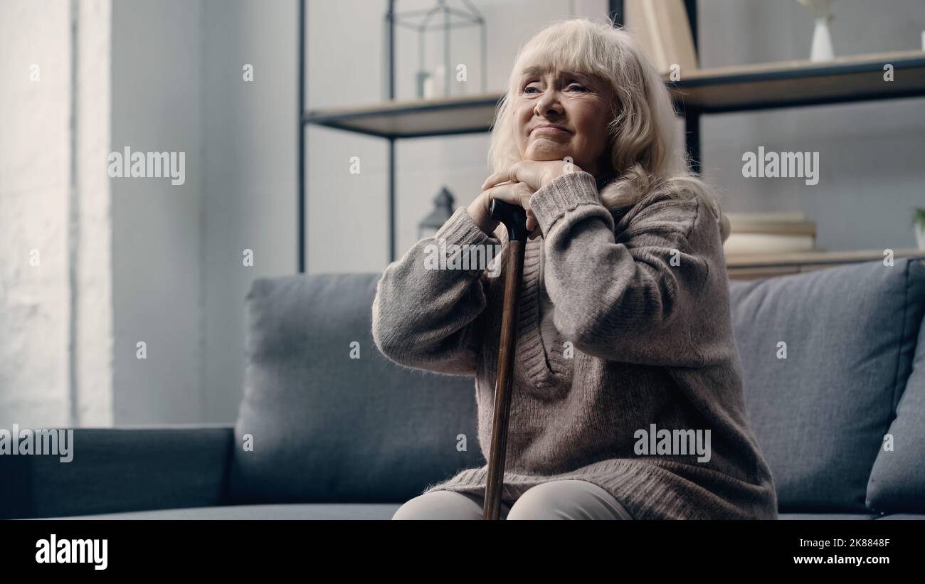 Sad Senior Woman With Dementia Sitting On Sofa While Leaning On Walking