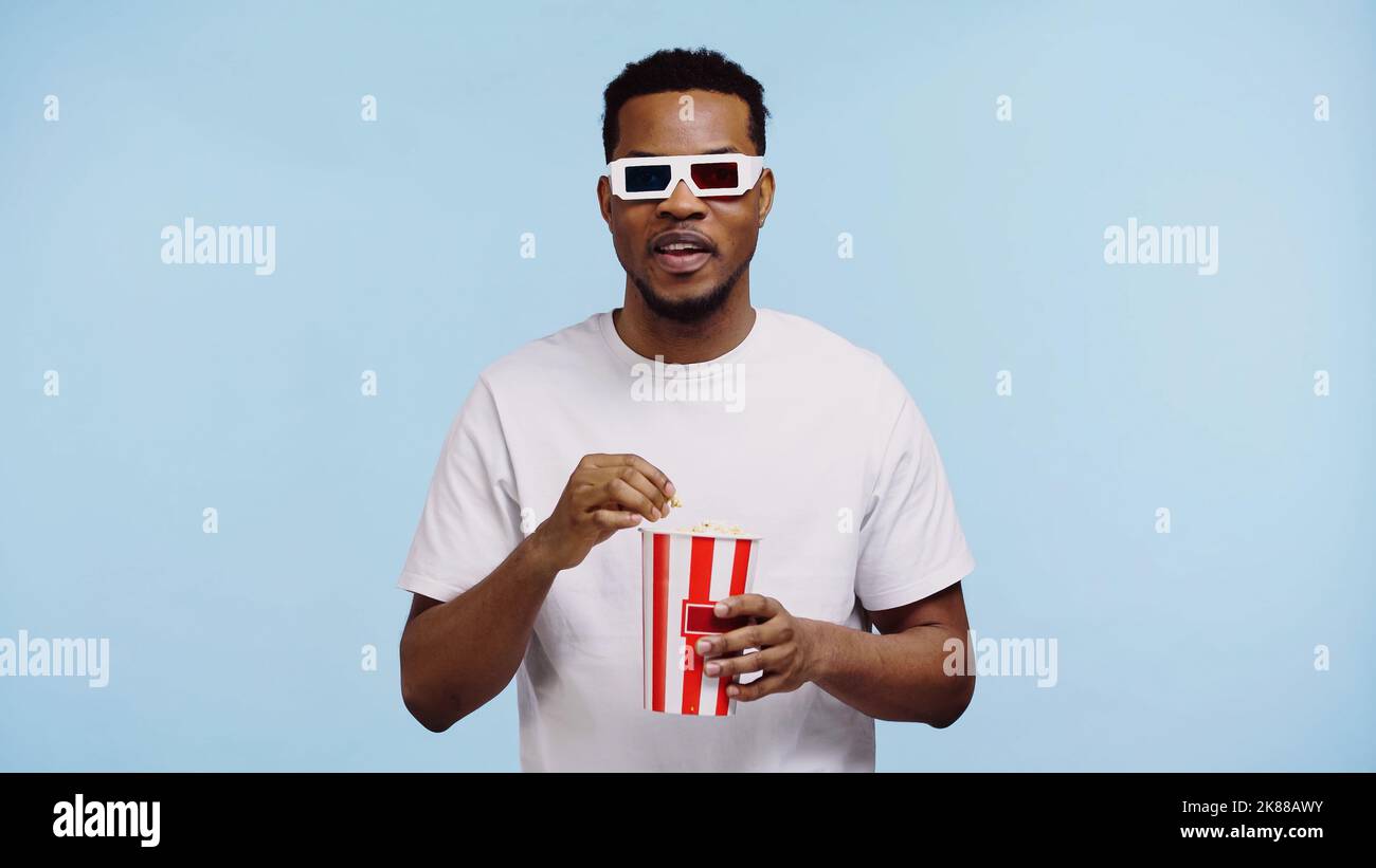 focused african american man in 3d glasses watching movie and holding popcorn isolated on blue ...