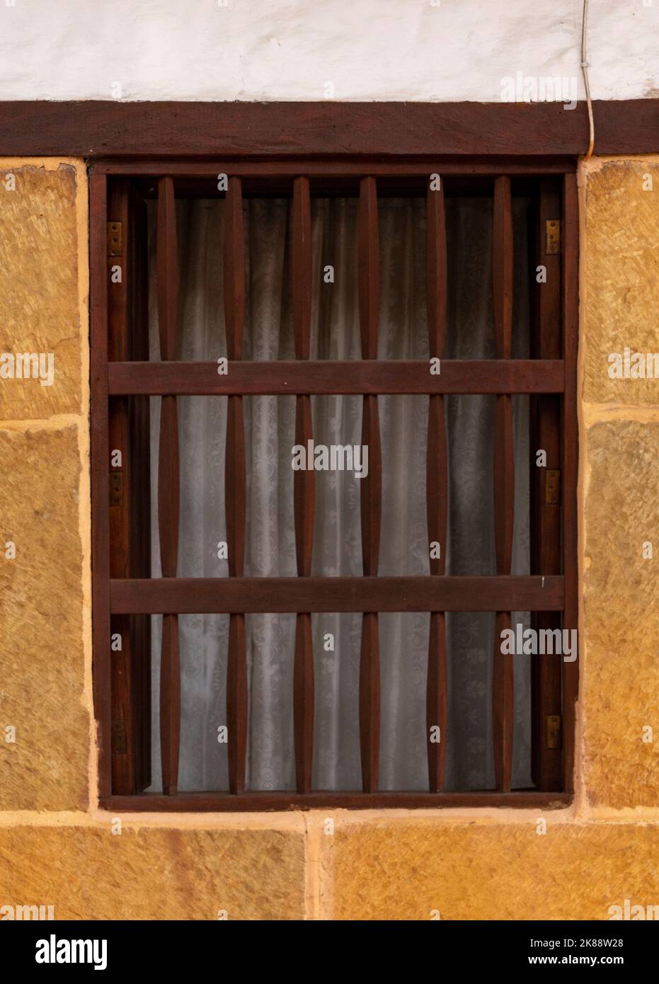 A window on a yellow stone wall, with a brown wooden grill and white ...