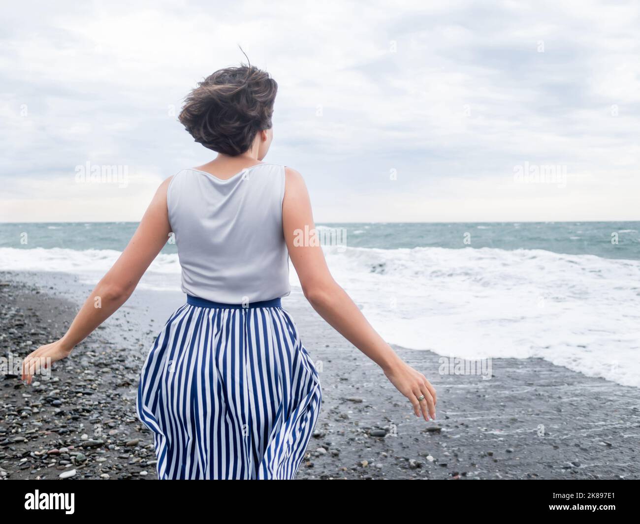 Happy woman greets stormy sea on pebble beach. Woman with hair ruffled ...