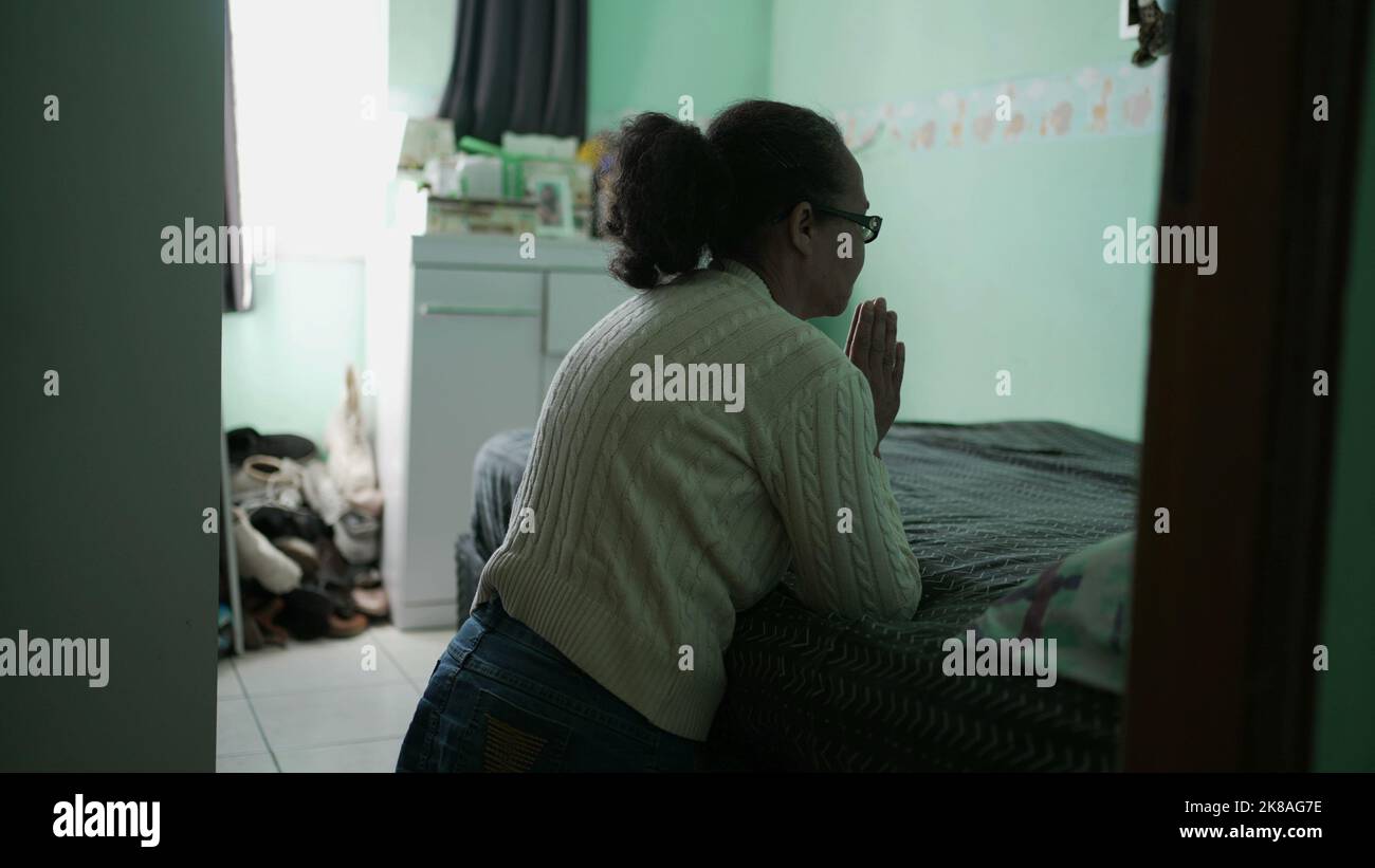 One older woman praying at home. A spiritual senior hispanic lady leaning by the bedside in ...
